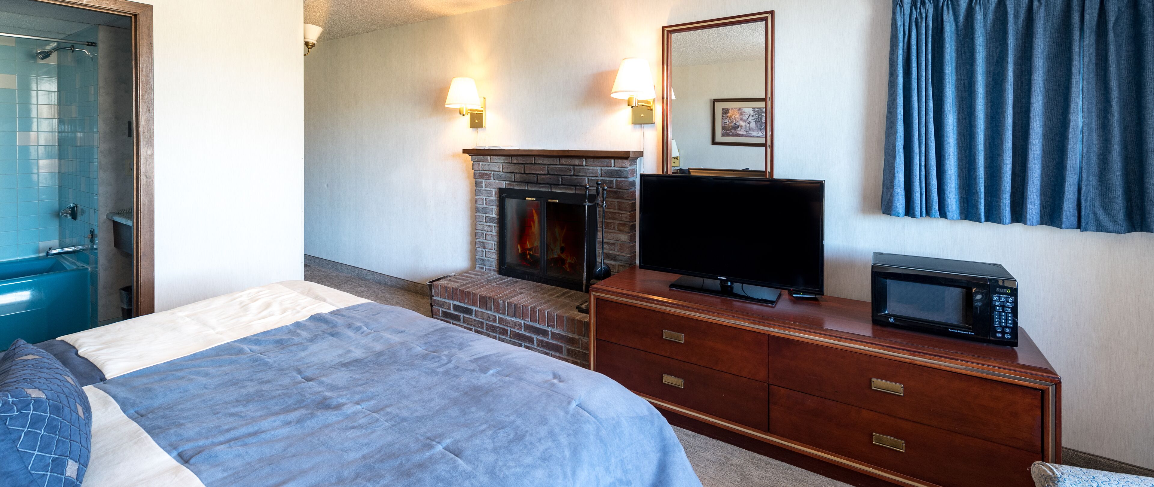 Quarto de hotel com cama, televisão, micro-ondas, lareira e banheiro com chuveiro azul.