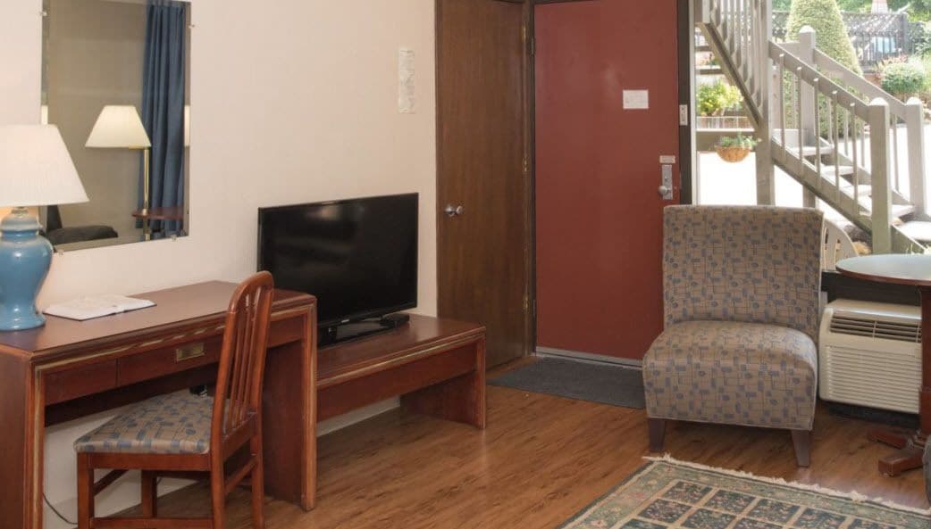 Quarto com escrivaninha de madeira, televisão, poltrona estampada e uma pequena mesa.