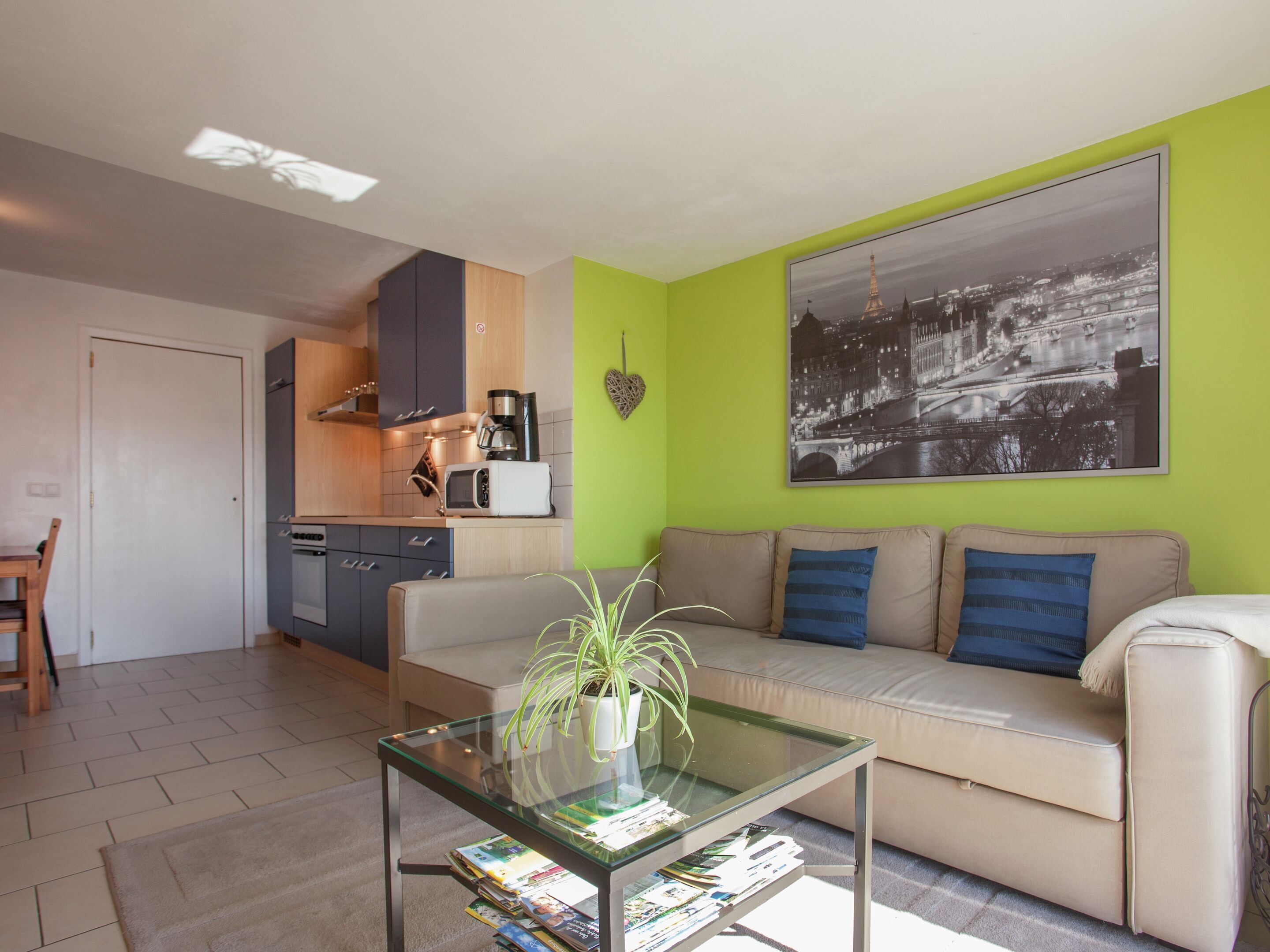 Uma sala de estar moderna com um sofá, uma mesa de centro de vidro com revistas, uma área de cozinha e uma pintura com vista para a cidade na parede.
