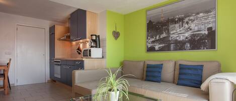 Uma sala de estar moderna com um sofá, uma mesa de centro de vidro com revistas, uma área de cozinha e uma pintura com vista para a cidade na parede.