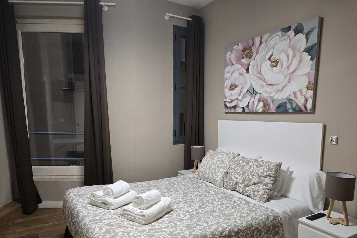 Uma cama bem arrumada com colcha estampada, duas luminárias de cabeceira, uma mesinha de cabeceira e um quadro de flores na parede.