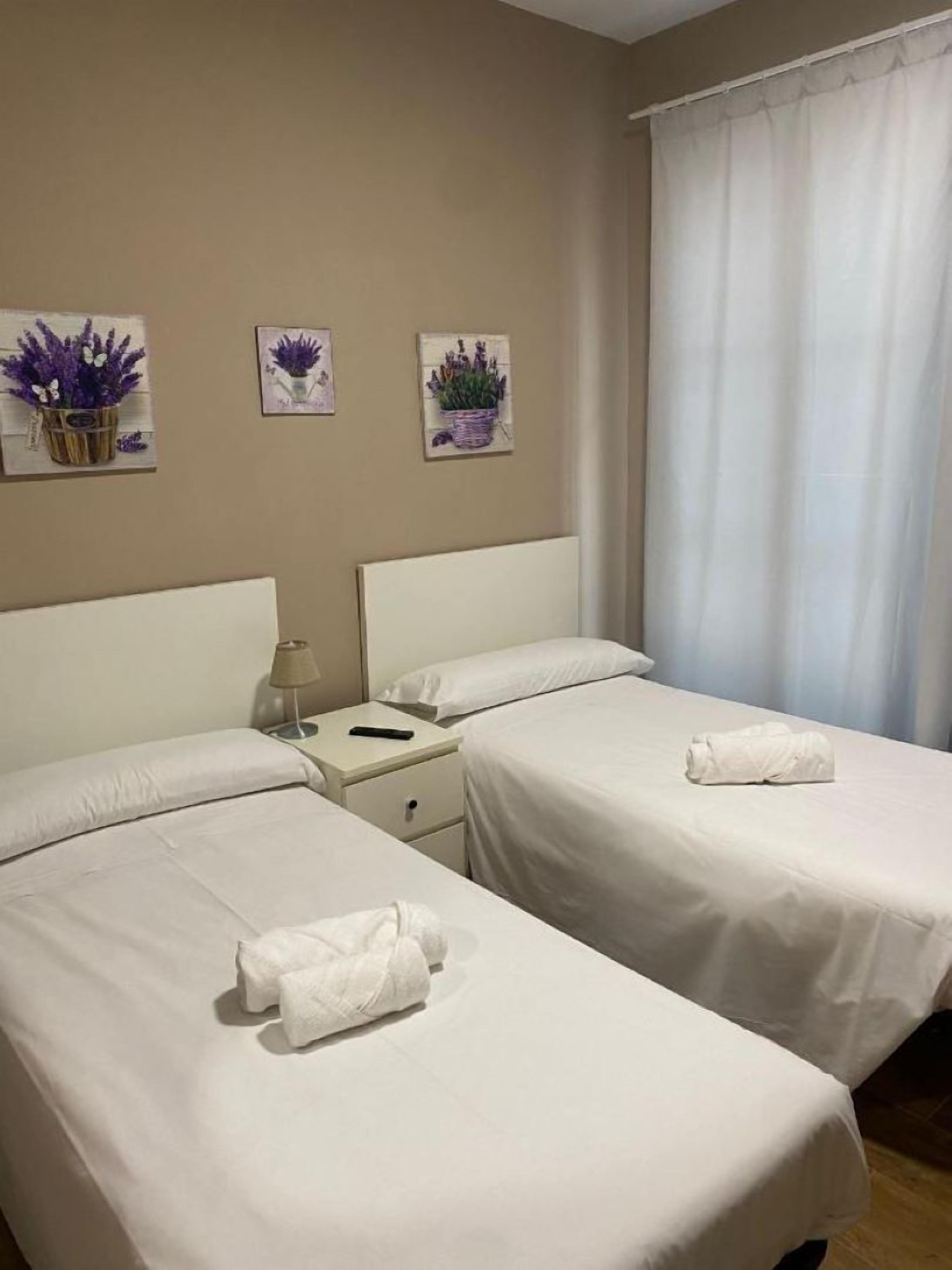 Quarto de hotel com duas camas, uma mesa de cabeceira, uma luminária e três quadros de flores fixados na parede.