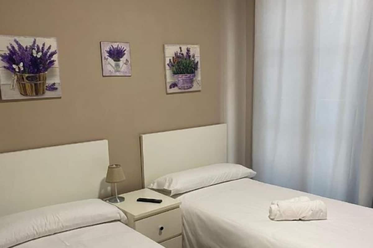 Quarto de hotel com duas camas, uma mesa de cabeceira, uma luminária e três quadros de flores fixados na parede.