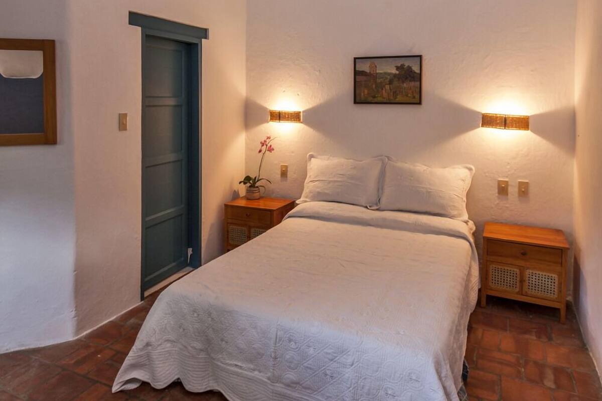 Quarto com cama, mesa de cabeceira de madeira, porta azul e um quadro pendurado na parede.