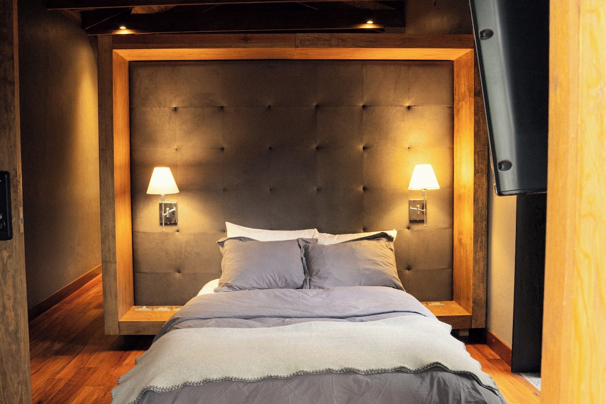 Quarto com cama grande, piso de madeira, cabeceira estofada e duas luminárias fixadas na parede.