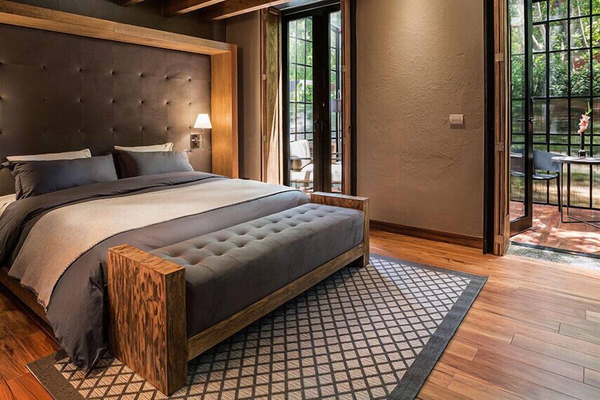 Quarto moderno com uma cama grande, cabeceira de madeira, duas luminárias de cabeceira e vista para um pátio externo.