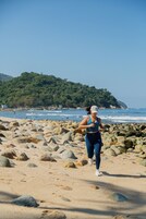 Uma pessoa corre numa praia rochosa, com uma colina arborizada ao fundo.