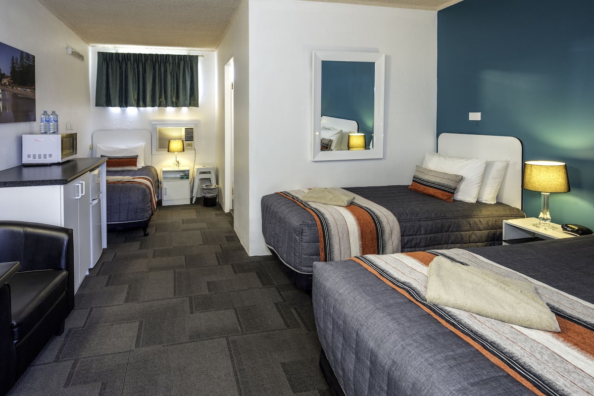 Quarto de hotel com duas camas, uma escrivaninha, uma cadeira, uma kitchenette compacta e um quadro na parede.