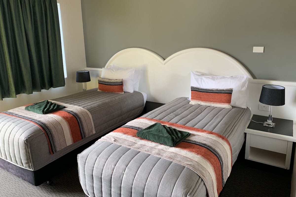 Quarto de hotel com duas camas, cada uma com colcha listrada e almofadas, uma mesa de cabeceira com abajur, e uma janela com cortinas verdes.