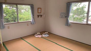 Quarto com piso de tatame, duas janelas com cortinas, ar-condicionado fixado na parede e uma pequena prateleira com um quadro emoldurado.