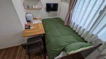 Quarto pequeno e bem arrumado, com escrivaninha de madeira, cama verde, cadeira, TV fixada na parede e janela com cortinas.