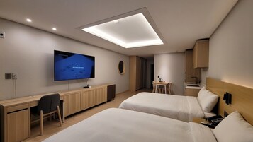Quarto de hotel moderno com duas camas, uma escrivaninha com cadeira, televisão, uma mesinha com cadeira e uma cozinha compacta.