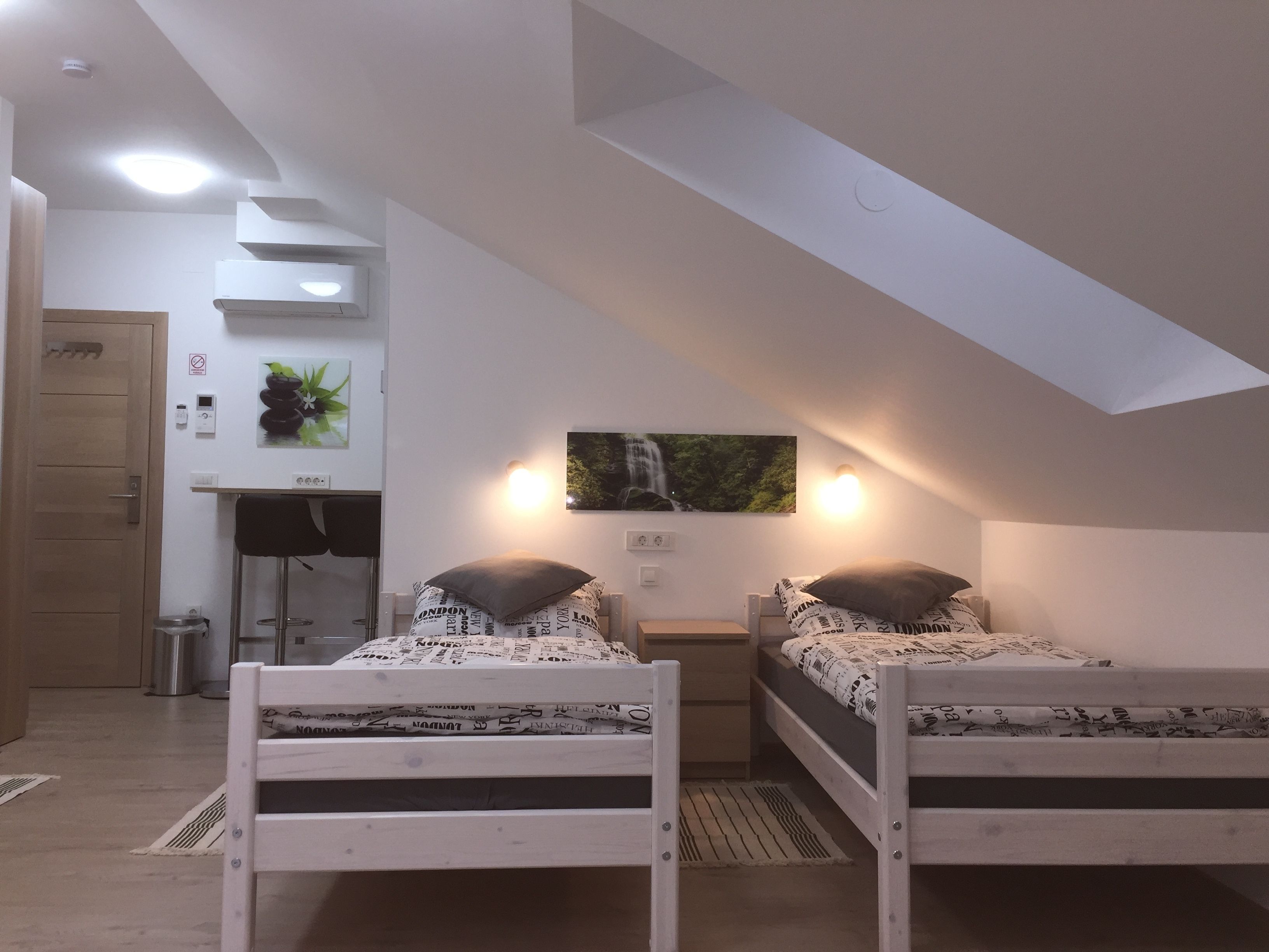 Quarto com duas camas de solteiro, escrivaninha, cadeira, mesa pequena, luz acoplada à parede, quadro de uma cascata e uma porta.