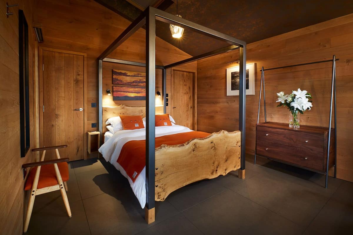 Uma cama de madeira com dossel, com colcha branca e laranja, um criado-mudo de madeira, uma cadeira e um aparador com um vaso de flores.