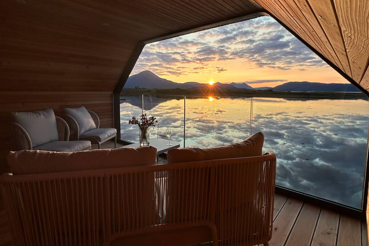 Um espaço de estar acolhedor com vista para um pôr do sol sobre montanhas e nuvens.