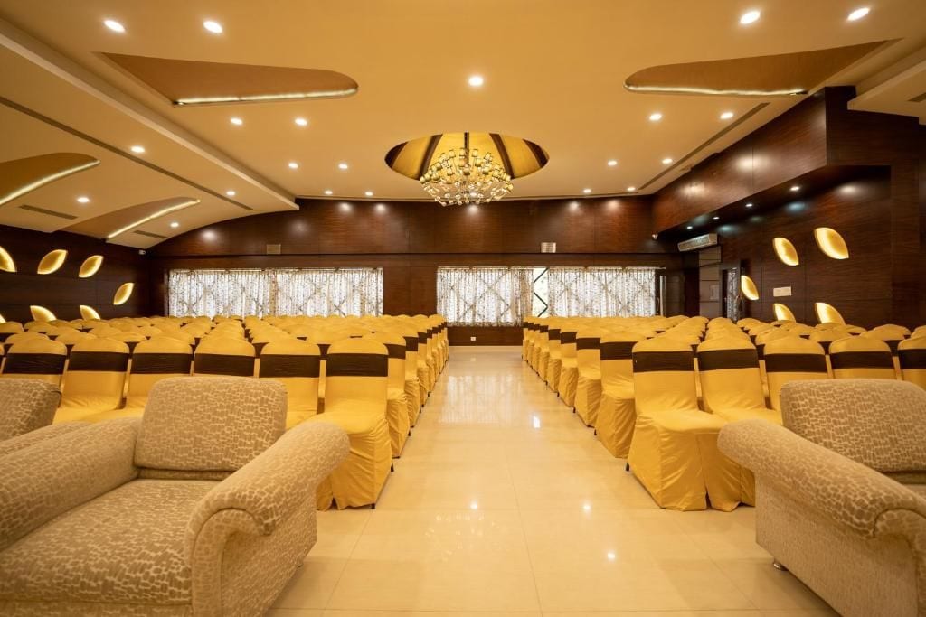 Uma sala de conferências com fileiras de cadeiras, um candelabro e uma janela grande com cortinas.