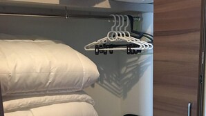 Quarto pequeno com uma cama, uma mesinha e uma cadeira. Há uma prateleira com almofadas e um rolo de papel higiênico.