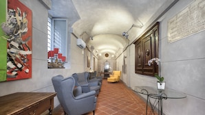 Um corredor com piso de azulejos, um banco de madeira, uma poltrona azul, uma poltrona amarela e uma mesa de vidro com um vaso de planta.