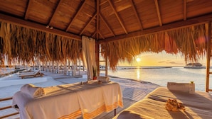 Um spa à beira-mar com um deck de madeira, mesas de massagem e vista para o pôr do sol sobre o oceano.
