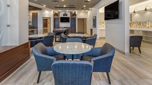 Um hall de hotel moderno com uma mesa de jantar redonda rodeada por cadeiras, uma área de cozinha ao fundo e uma televisão de tela plana na parede.