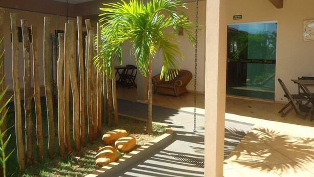 Uma área de varanda coberta com uma palmeira, cerca de madeira e um espaço de estar com mesa e cadeiras.