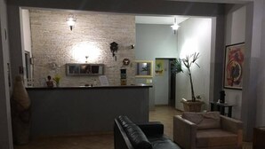 Sala de estar com sofá de couro preto, poltrona bege e mesa de centro. Há uma área de cozinha com parede de tijolos e balcão. Um vaso com planta é visível no canto.