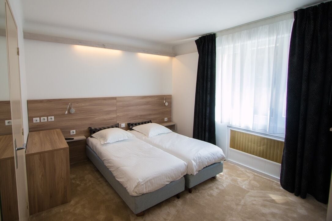 Quarto de hotel com duas camas, cabeceira de madeira, mesa de cabeceira, guarda-roupa e uma janela grande com cortinas.