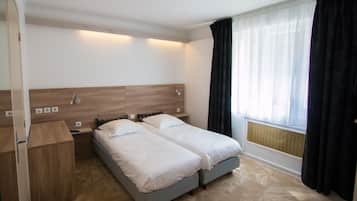 Quarto de hotel com duas camas, cabeceira de madeira, mesa de cabeceira, guarda-roupa e uma janela grande com cortinas.