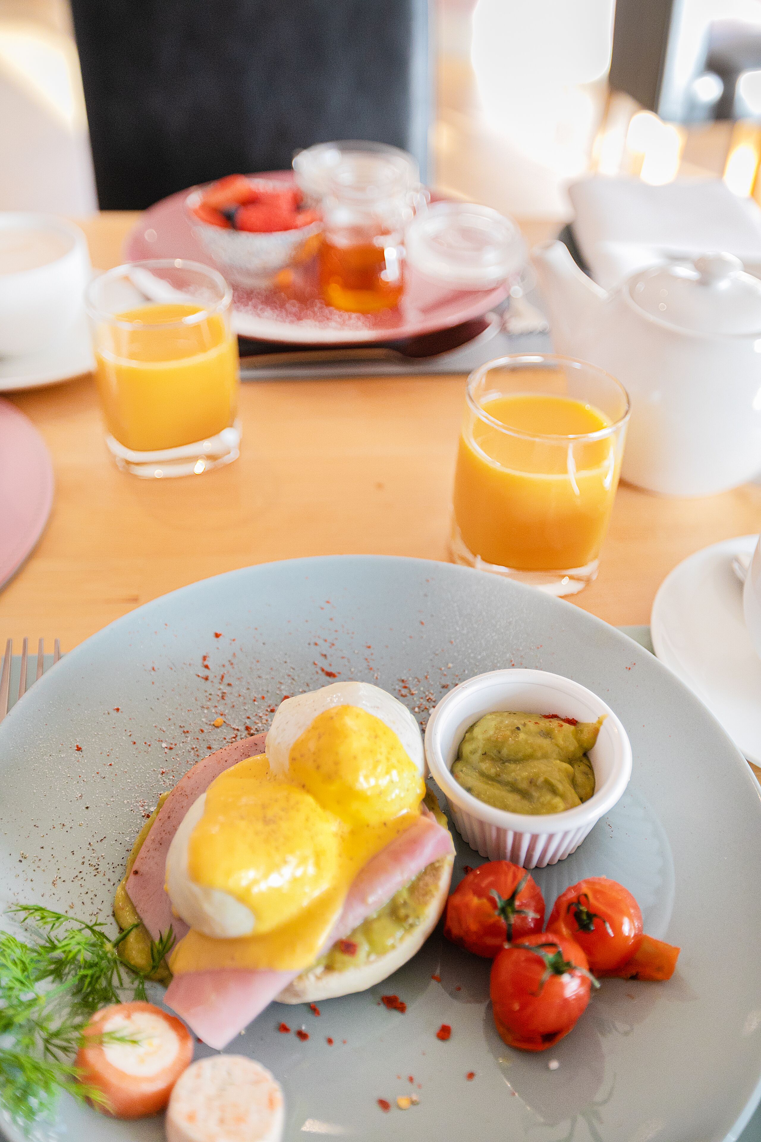 Prato de café da manhã com ovos, presunto e tomates, acompanhado de um copo de suco de laranja e uma pequena tigela de molho.
