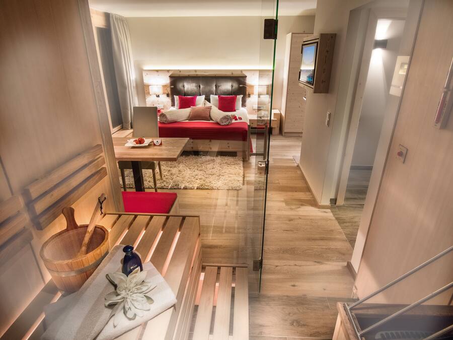 Quarto de hotel moderno com piso de madeira, cama com roupa de cama vermelha e branca, uma pequena mesa com uma garrafa e flores, e uma TV fixada na parede.