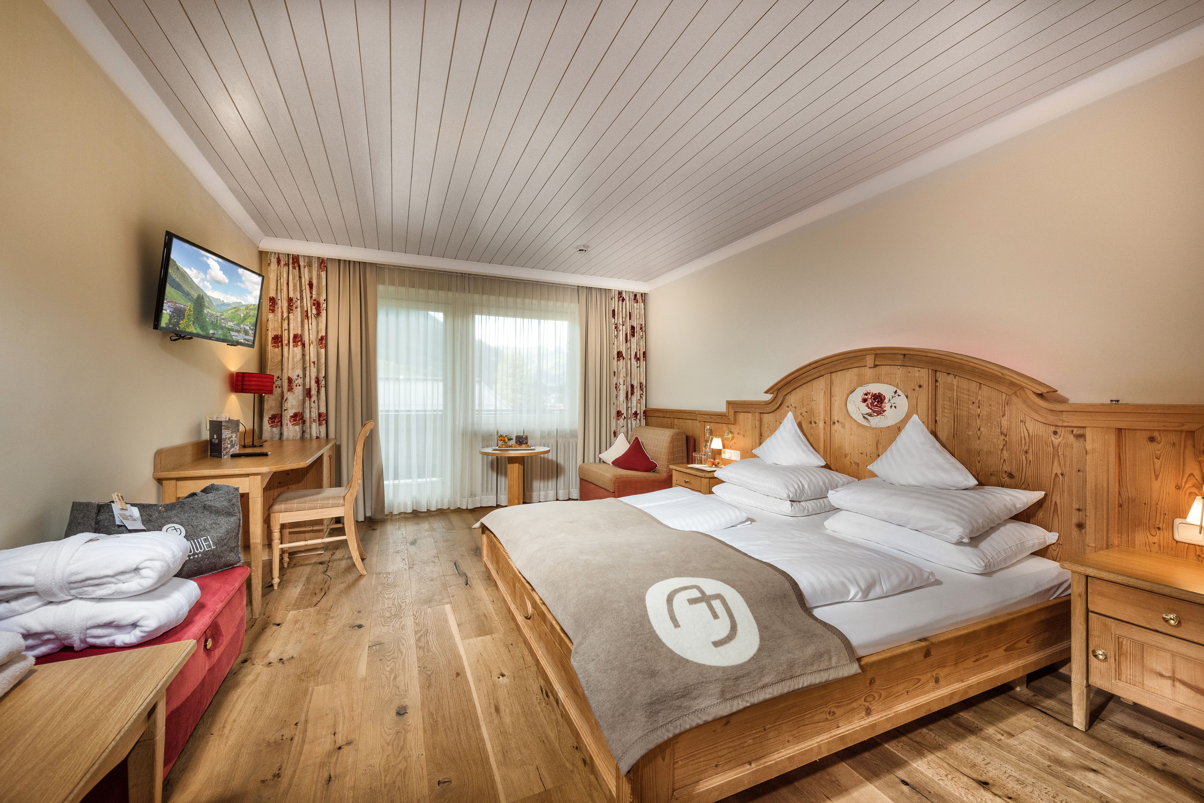 Quarto de hotel com uma cama de madeira, uma escrivaninha, uma cadeira, uma televisão e uma mesinha de cabeceira com abajur.