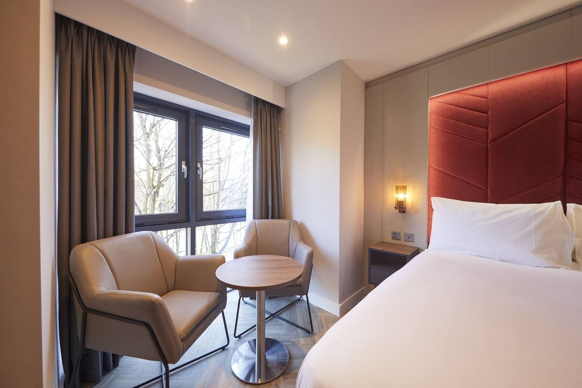 Quarto de hotel com uma cama, uma mesa redonda, duas cadeiras, uma janela com cortinas e cabeceira vermelha.