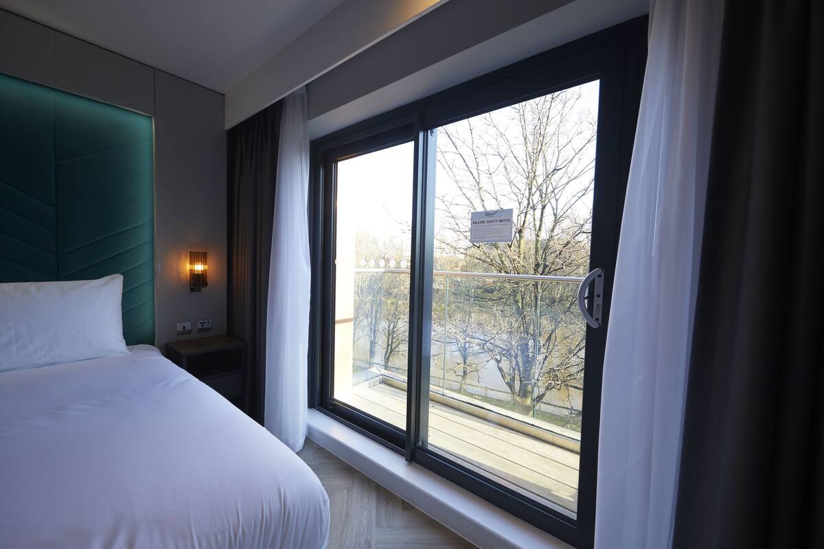 Quarto de hotel com uma cama grande, mesa de cabeceira com abajur e janela com vista para árvores e um letreiro.
