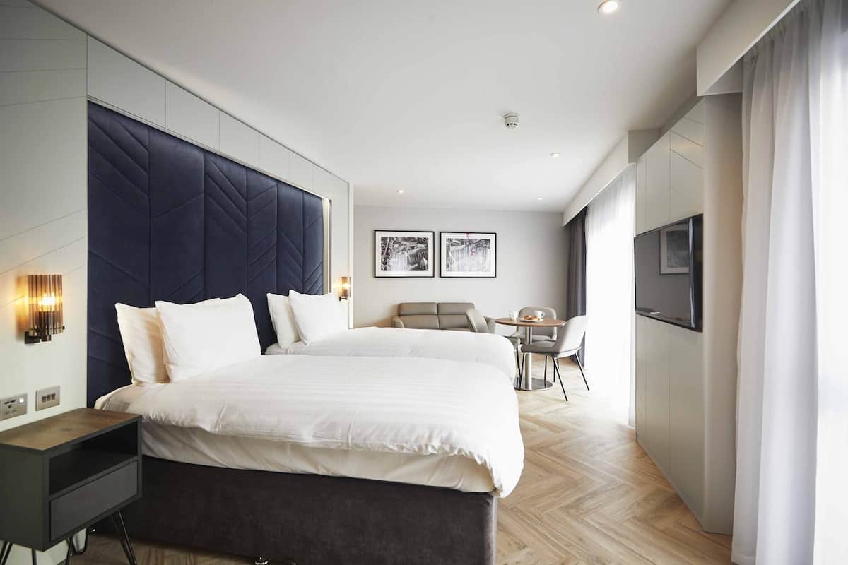 Quarto de hotel moderno com uma cama grande, mesas de cabeceira, um espaço de estar com mesa e cadeiras e uma TV fixada na parede.