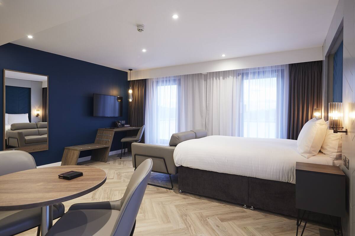 Quarto de hotel moderno com uma cama grande, uma escrivaninha, uma cadeira, uma mesa redonda e uma televisão.