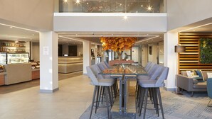 Uma área de refeições moderna com mesa de vidro e banquetas de bar, um arranjo central com uma árvore decorativa e um teto alto com iluminação embutida.