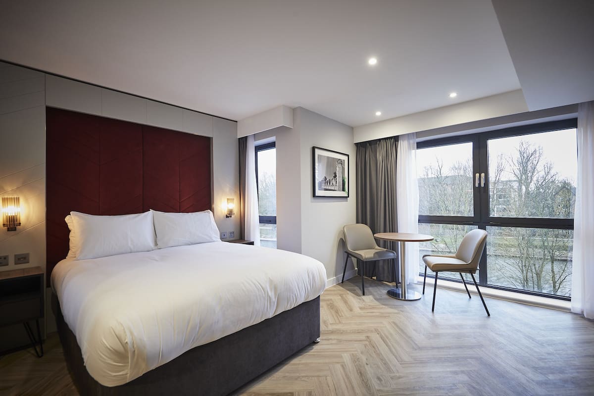 Quarto de hotel moderno com uma cama grande, uma escrivaninha e uma cadeira, uma janela com vista para árvores e um quadro na parede.
