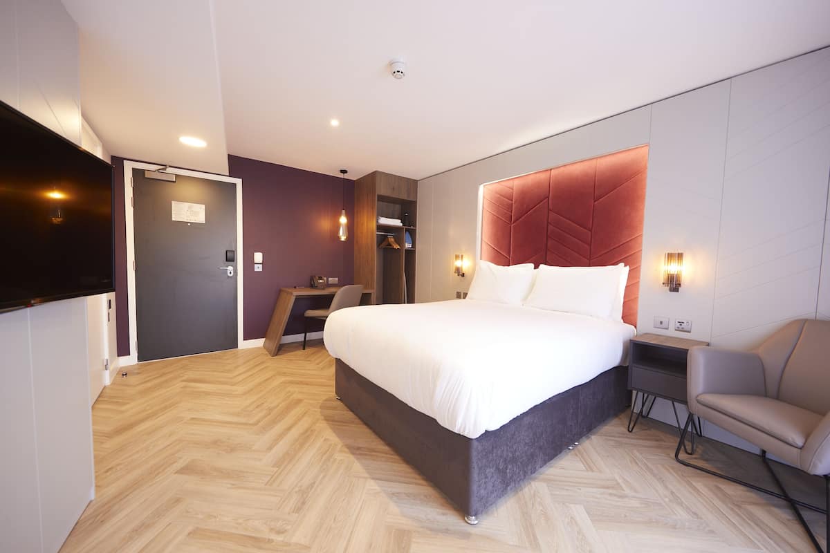 Quarto de hotel moderno com uma cama grande, escrivaninha, cadeira e televisão na parede.
