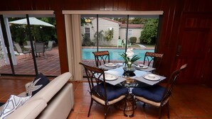 Uma sala de jantar com uma mesa posta para quatro pessoas, rodeada de cadeiras, e vista para uma piscina e uma casa ao fundo.
