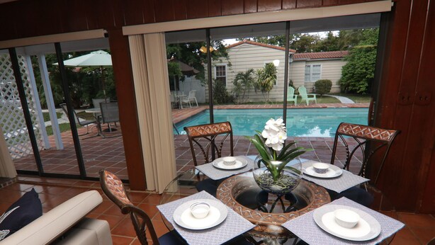 Uma área de refeições com mesa e cadeiras de vidro, vista para a piscina e um pátio com mobiliário externo.