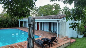Piscina com deck ladrilhado, área de varanda coberta com cadeiras e um edifício branco com telhado ladrilhado.