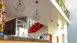 Uma área de recepção moderna com balcão de ripas de madeira, iluminação pendente, escada com guarda-corpo vermelho e varanda com vegetação.