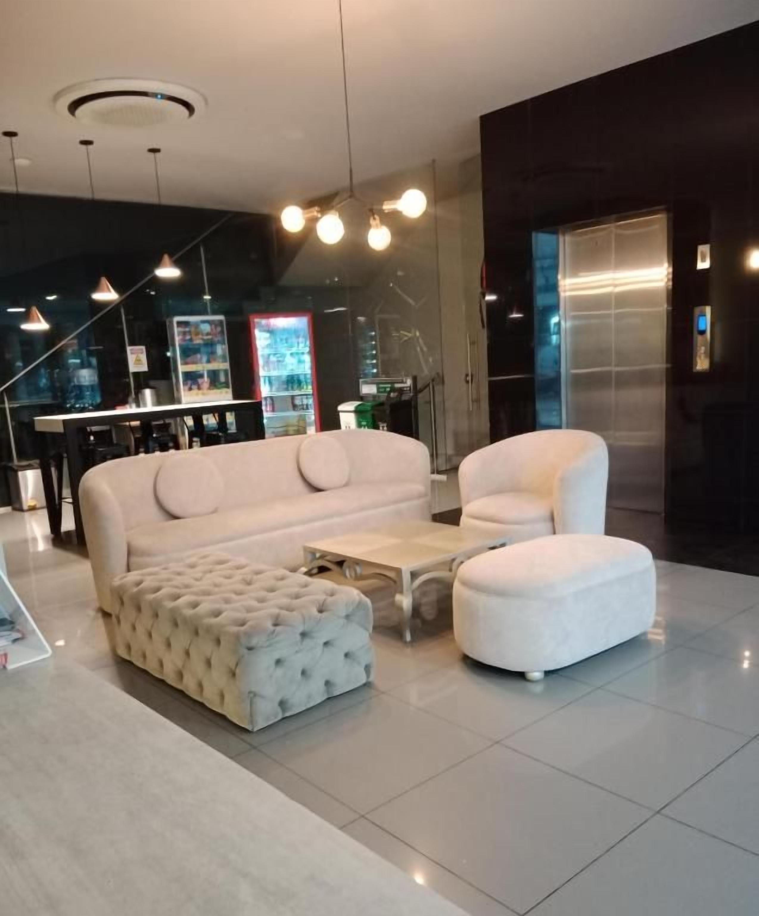 Um lobby de hotel moderno com sofá, banqueta e poltrona, uma mesa de centro e um bar ao fundo.