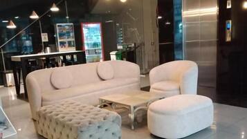 Um lobby de hotel moderno com sofá, banqueta e poltrona, uma mesa de centro e um bar ao fundo.