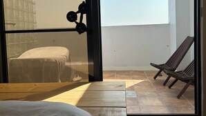 Quarto com cama, porta de correr de vidro que dá para uma varanda com cadeira e vista do céu.