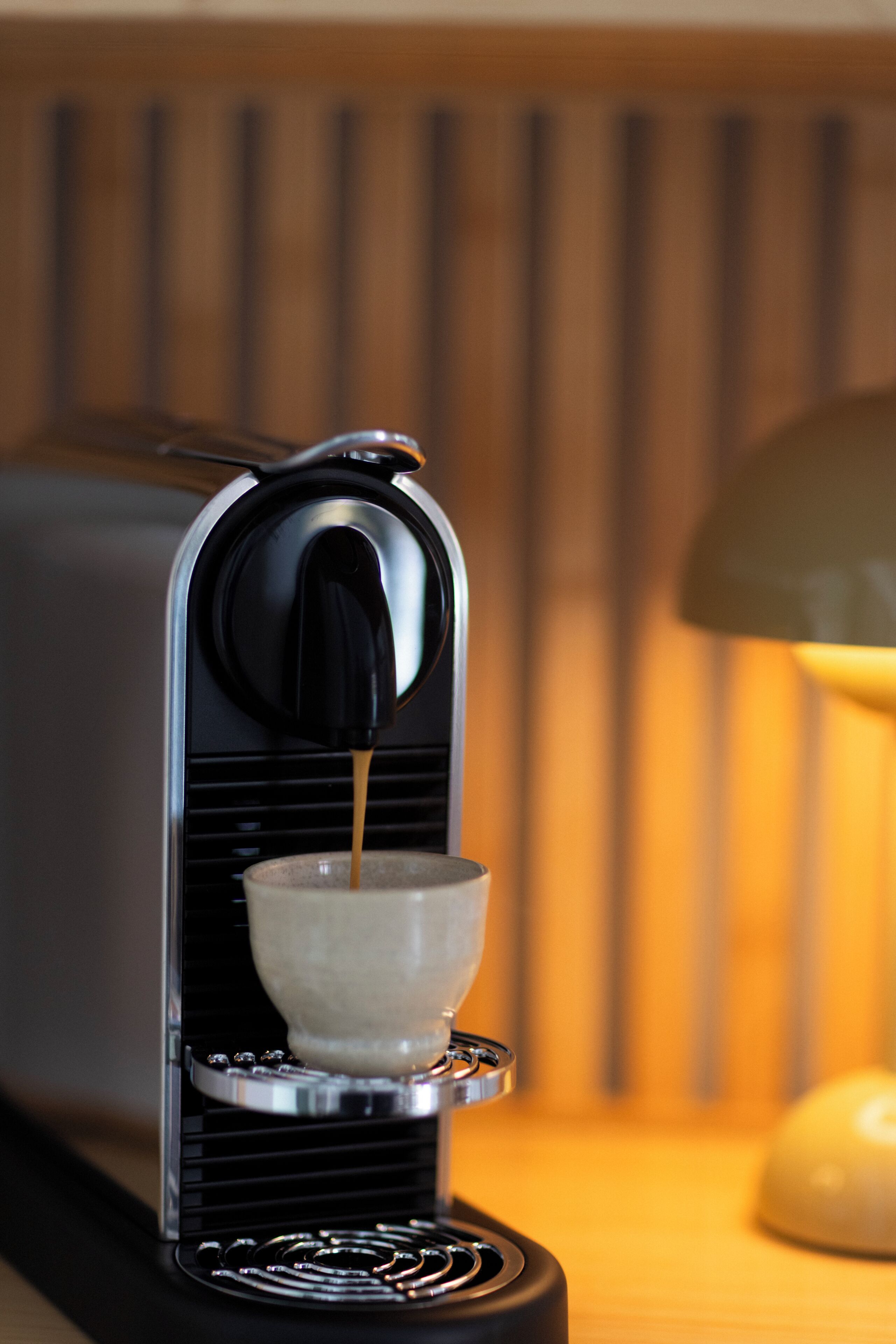 Máquina de café com uma xícara embaixo, uma luminária e fundo de madeira.