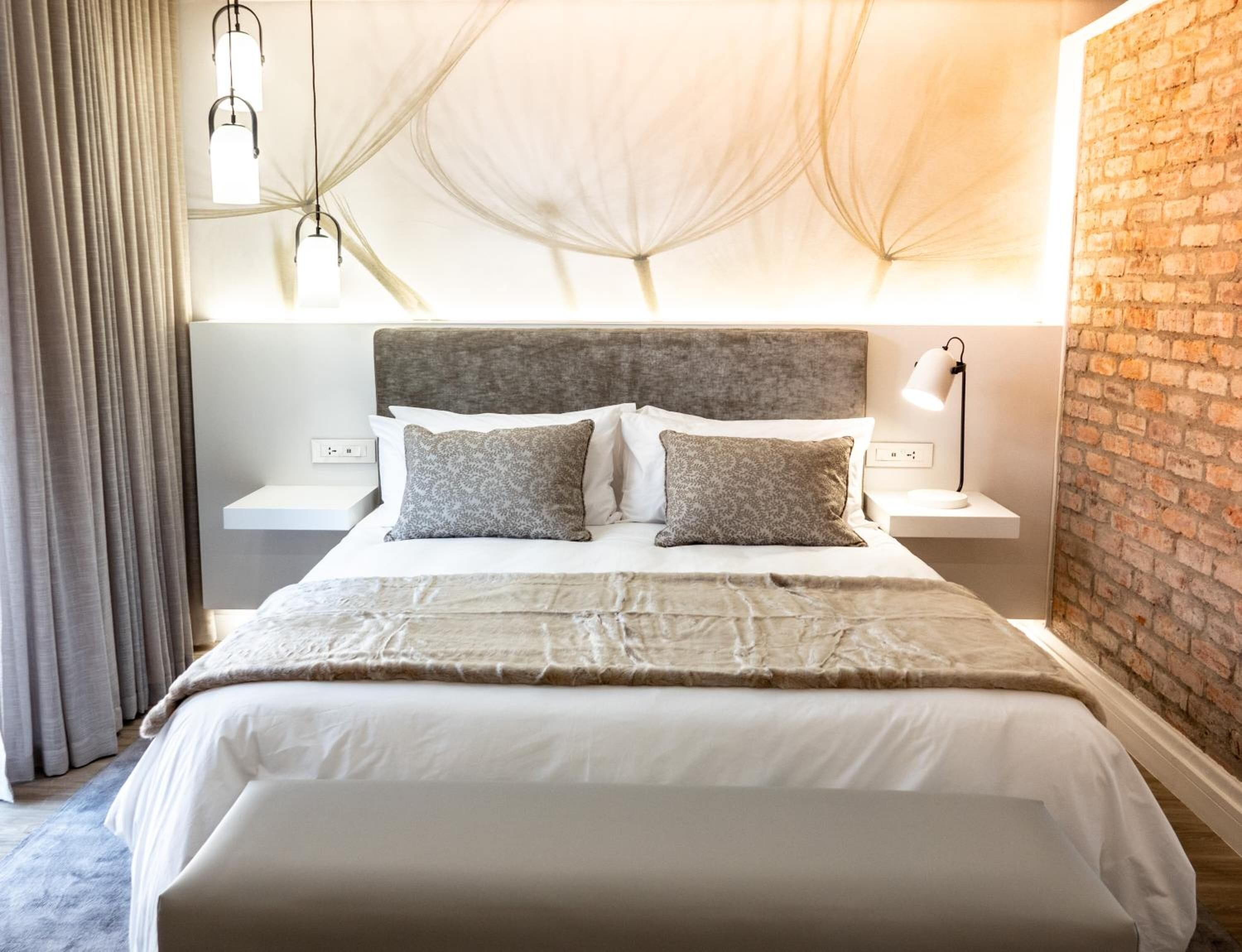 Quarto de hotel moderno com uma cama grande, duas mesas de cabeceira, parede de destaque em tijolo e um design decorativo no teto.