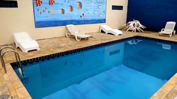 Piscina interna com piso ladrilhado, cadeiras de lounge brancas e um mural de peixes na parede.