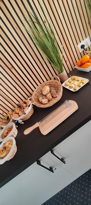 Uma mesa com diversos alimentos, incluindo pão, doces e laranjas, com um painel de ripas de madeira ao fundo.