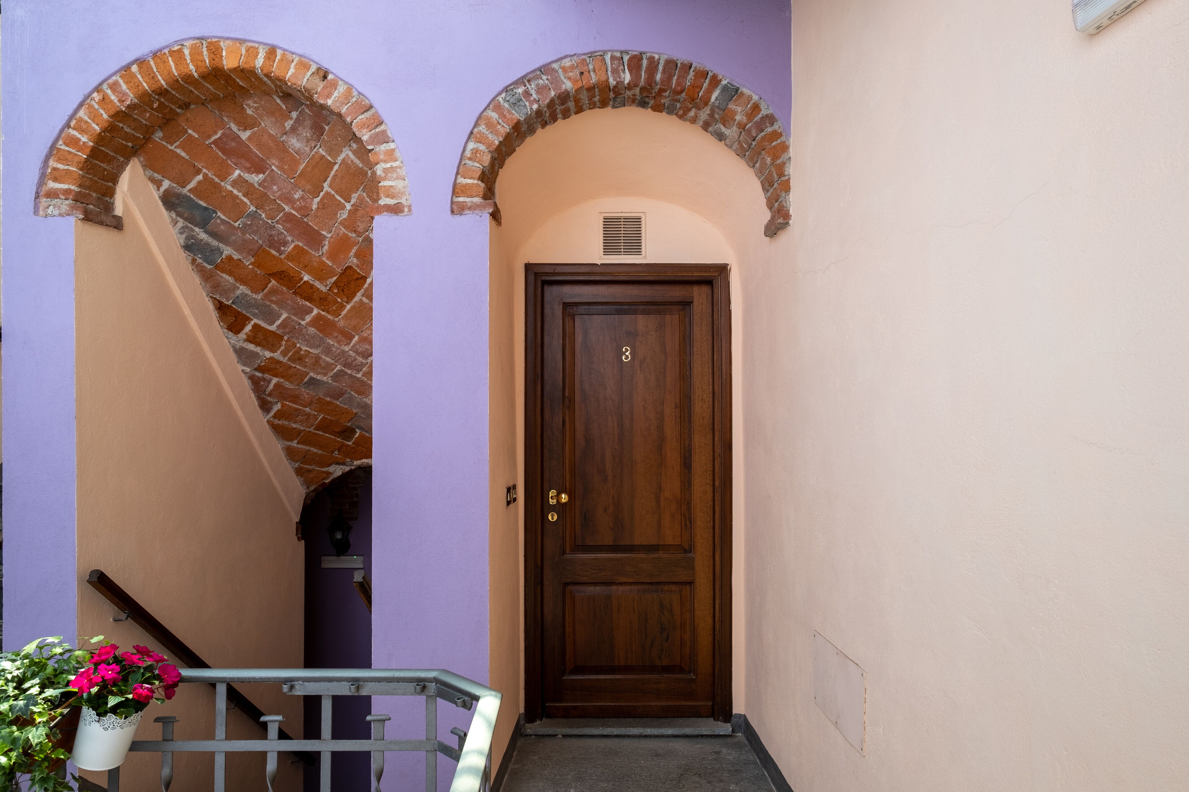 Um corredor estreito com uma porta de madeira marcada com o número 3, um arco de tijolos e um corrimão de metal com uma planta em vaso.
